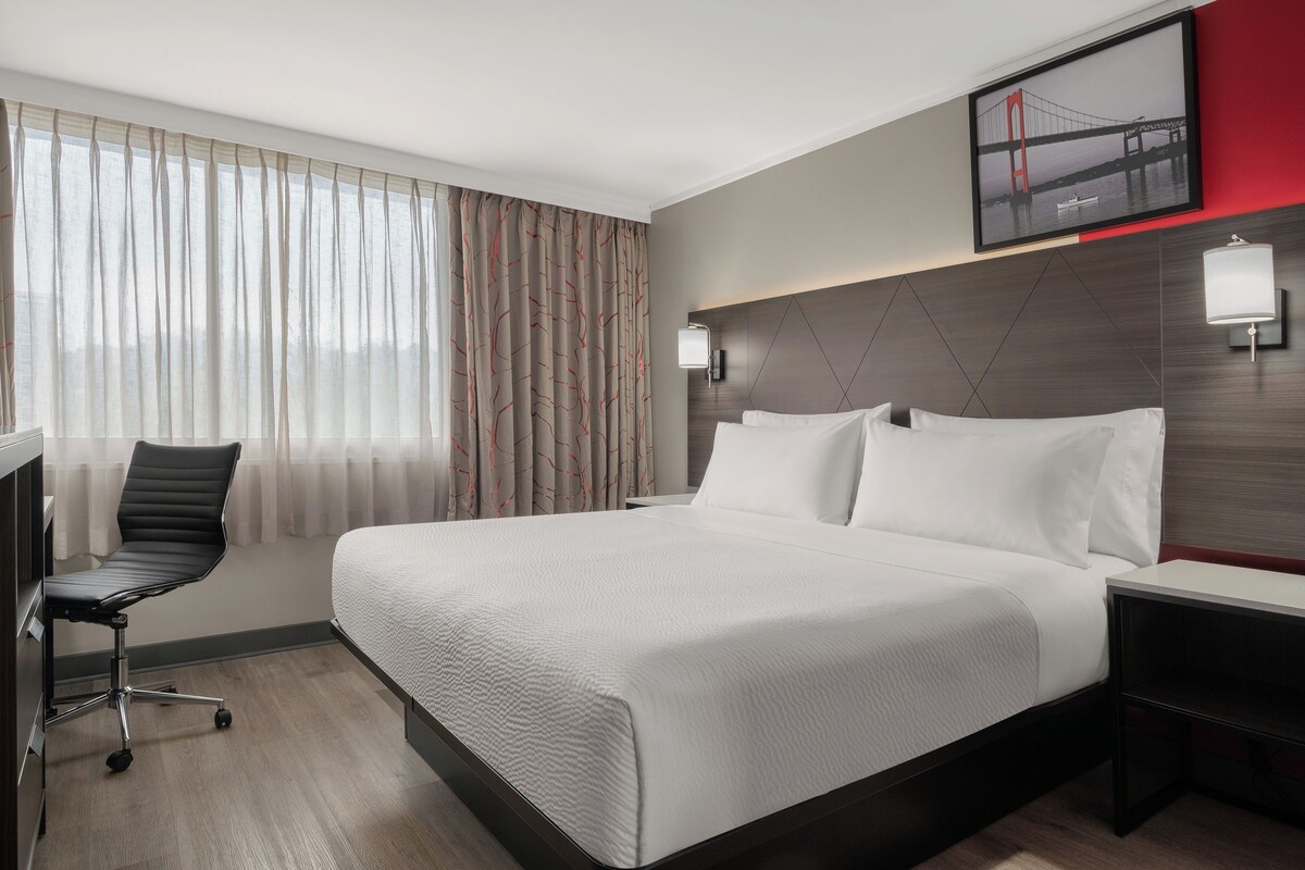 Quarto de hotel com cama, mesa de trabalho, cadeira e televisão fixada na parede.
