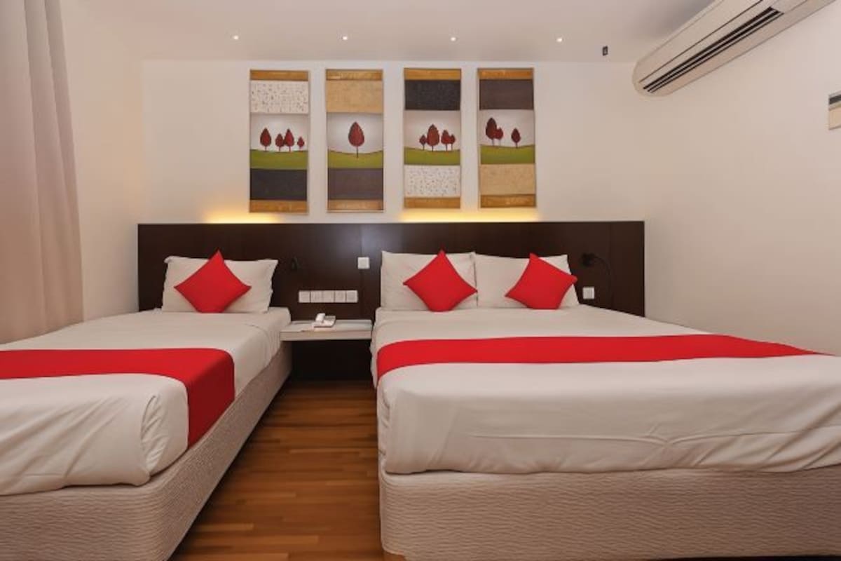 Quarto de hotel com duas camas, cada uma com roupa de cama branca e detalhes vermelhos, uma mesa de cabeceira e um ar condicionado fixado na parede.