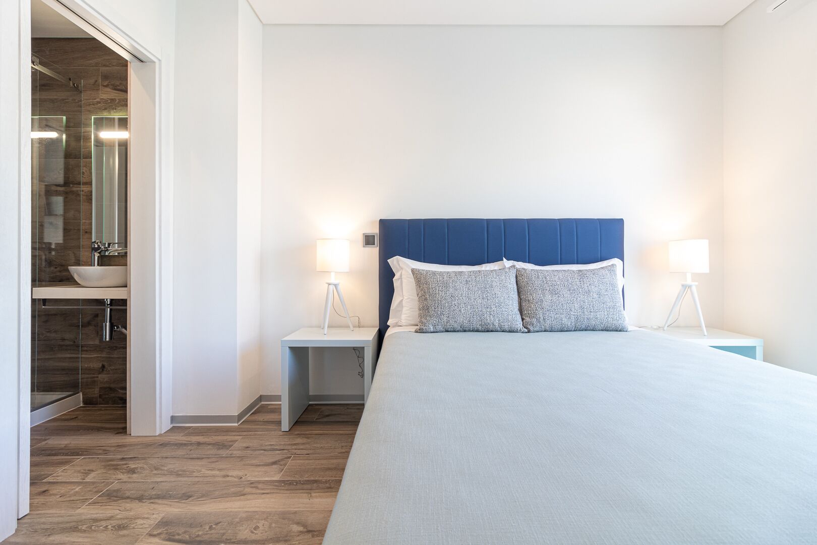 Quarto moderno com uma cama grande, duas mesas de cabeceira com luminárias, banheiro com box de vidro e piso de madeira.
