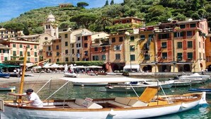 Uma marina com barcos atracados, um calçadão à beira-mar e edifícios coloridos ao fundo.