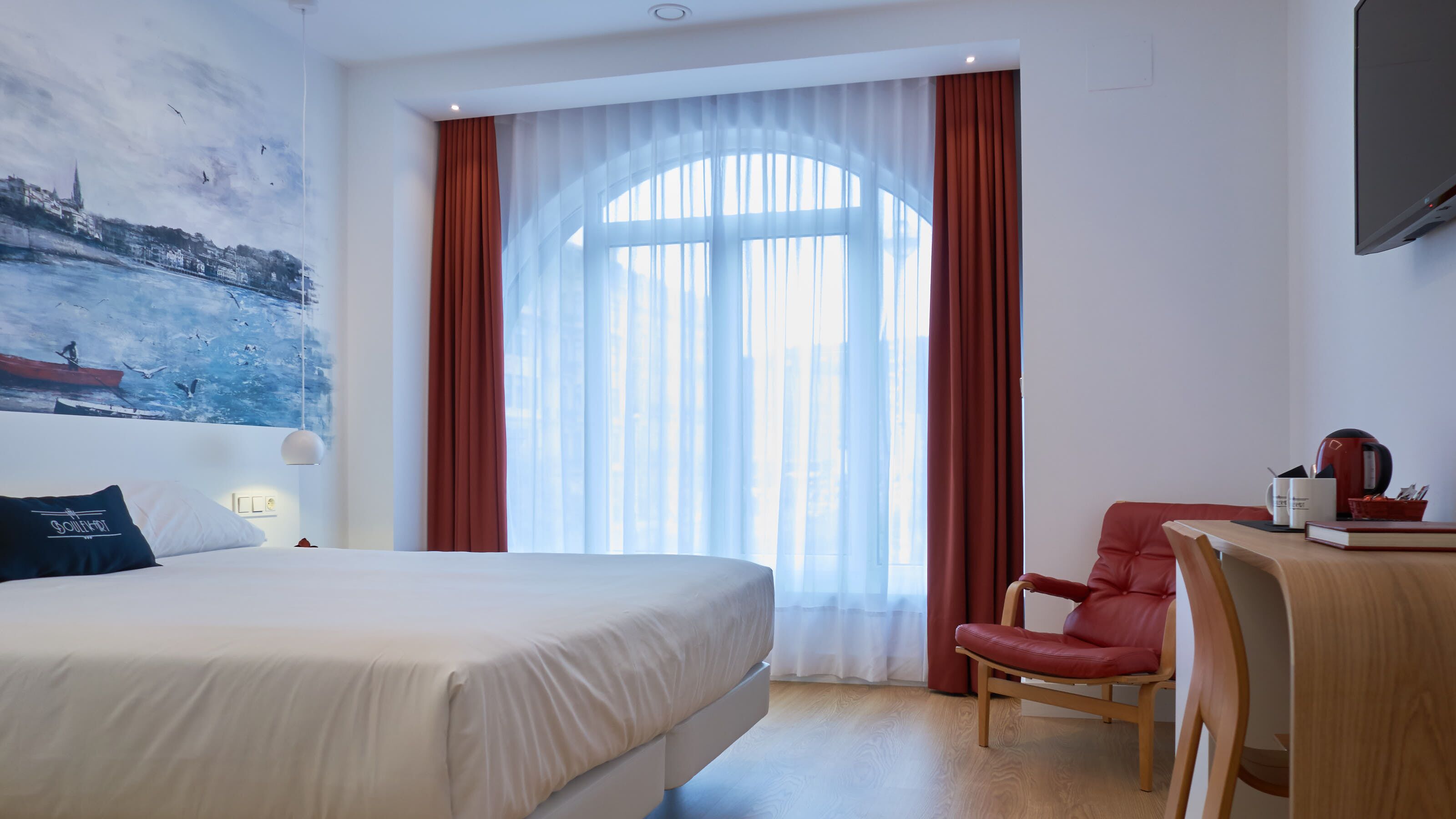 Quarto de hotel com cama, escrivaninha, cadeira, pintura e janela com cortinas.