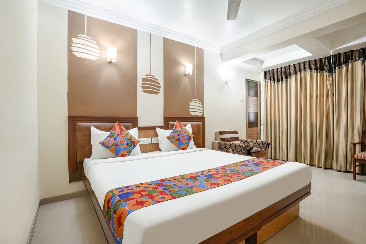 Quarto de hotel com uma cama grande, duas luzes pendentes, cabeceira de madeira, manta estampada e vista para o exterior através de uma janela com cortinas.