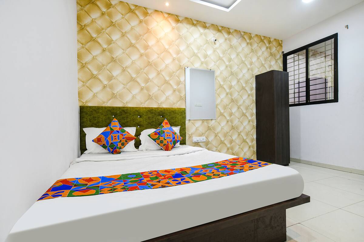 Cama bem arrumada com colcha geométrica colorida, cabeceira estofada e parede branca com janela.