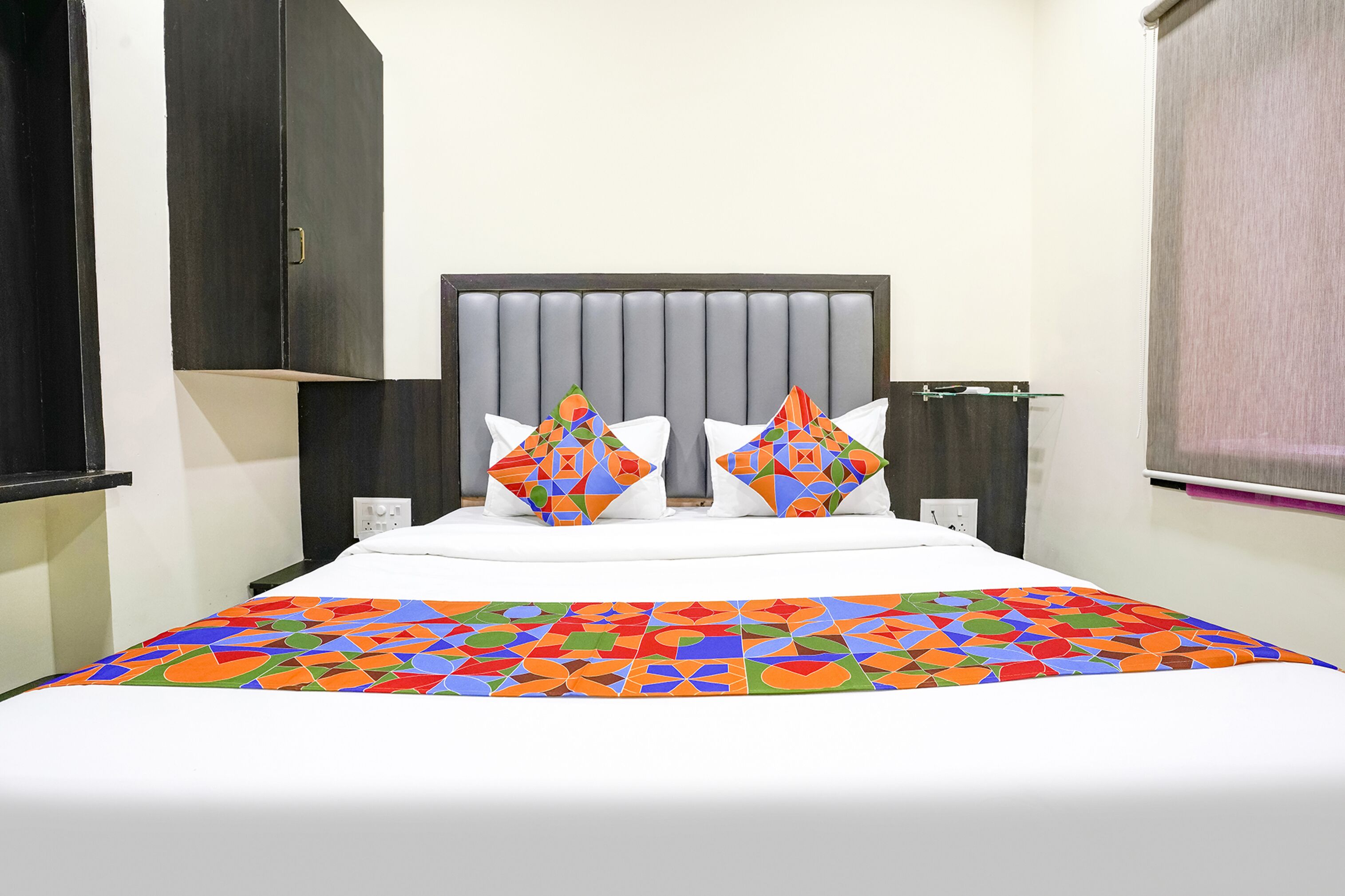 Uma cama com colcha de padrão geométrico colorido, duas almofadas com desenho semelhante, um painel de cabeceira estofado cinza e um guarda-roupa de madeira escura.