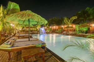 Área de piscina à noite, com espreguiçadeiras, guarda-sóis e um elemento aquático.