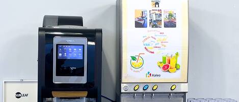 Uma máquina de café com visor digital e dispensador de bebidas, um quadro de menu fixado na parede com imagens e texto, e uma bandeja de copos plásticos.