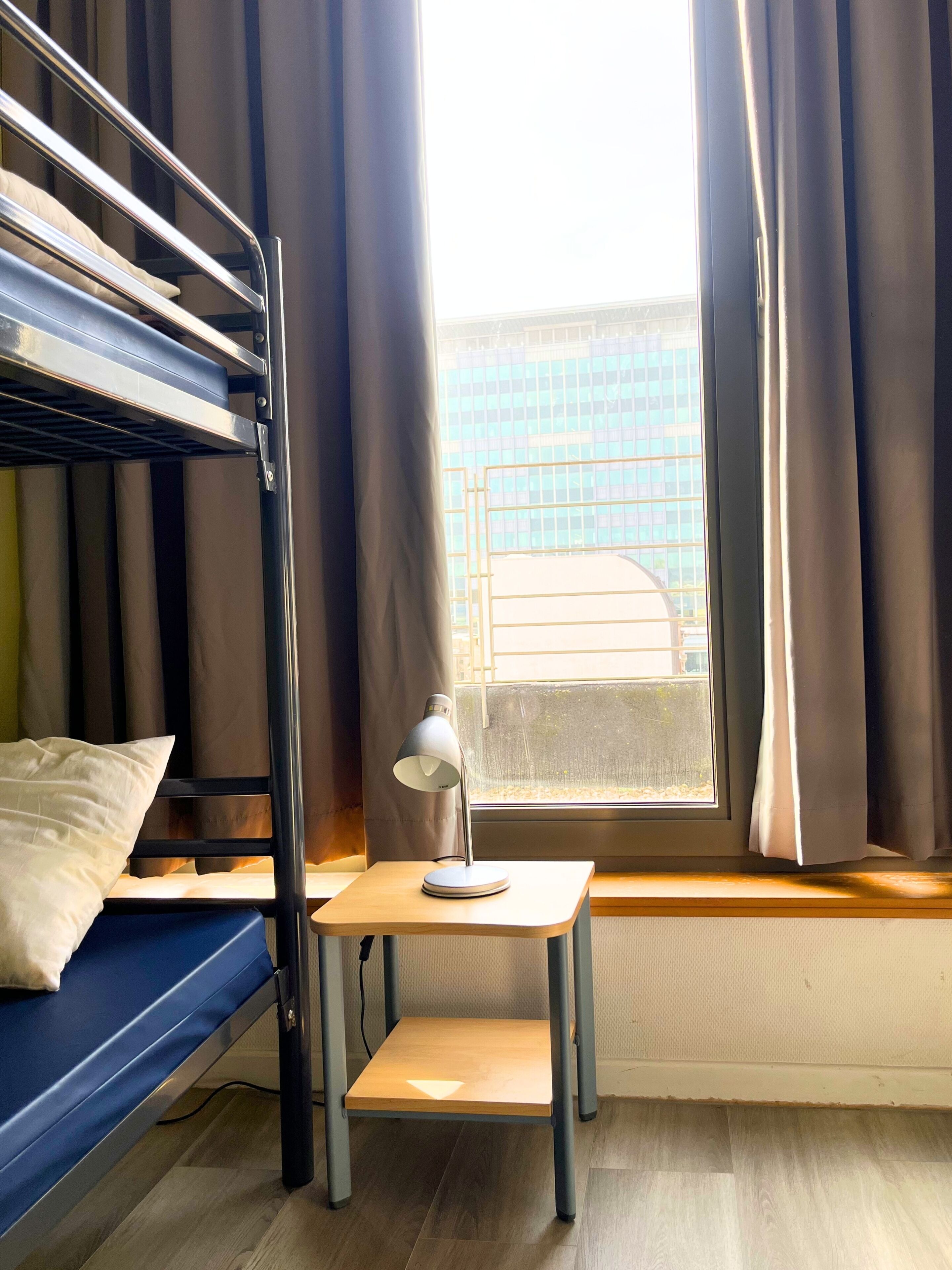 Uma cama beliche com escrivaninha e luminária, janela com cortinas e vista para um edifício.