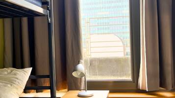 Uma cama beliche com escrivaninha e luminária, janela com cortinas e vista para um edifício.