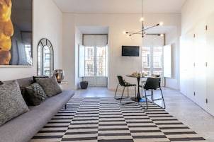 Uma sala de estar moderna com sofá, mesa de jantar com cadeiras, tapete listrado em preto e branco e TV fixada na parede.
