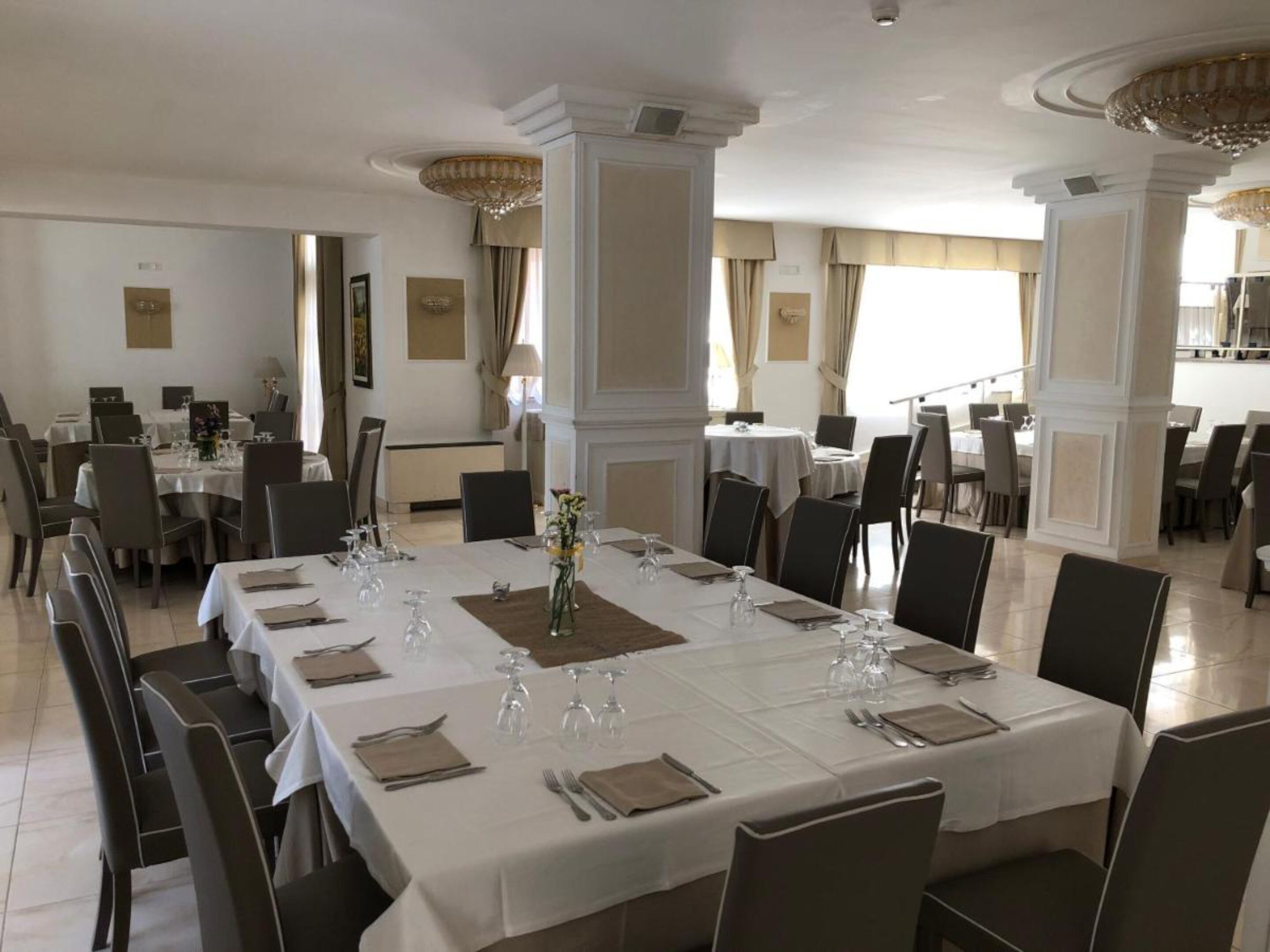 Uma sala de jantar formal com várias mesas preparadas para o serviço, elegantes lustres e um design arquitetônico clássico.