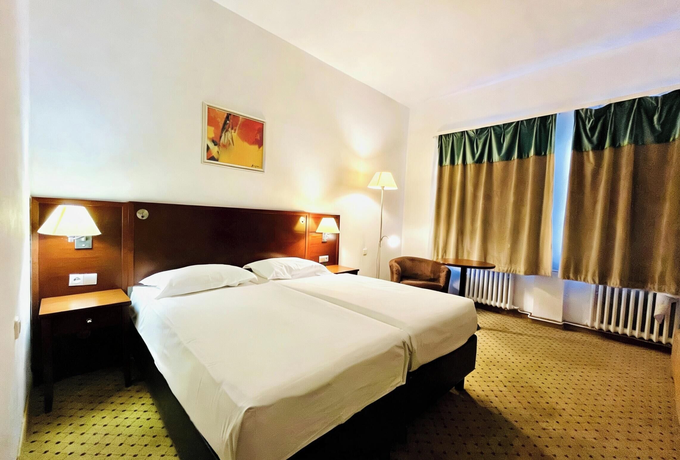 Quarto de hotel com uma cama de casal, duas luminárias de cabeceira, uma cabeceira, uma cadeira, um radiador e uma janela com cortinas.