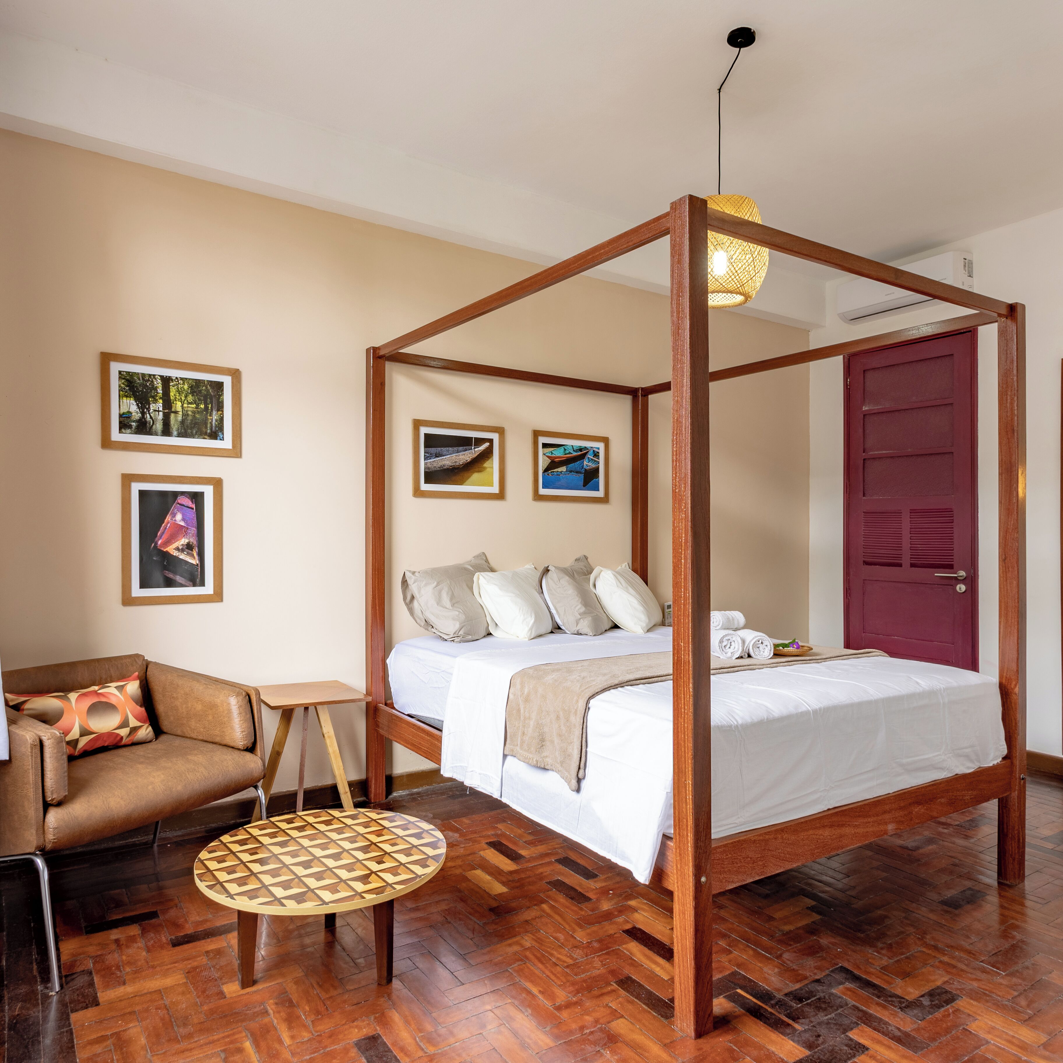 Uma cama com dossel e estrutura de madeira, uma mesa lateral redonda trançada, um sofá de couro marrom e uma porta vermelha em um quarto com piso de madeira.