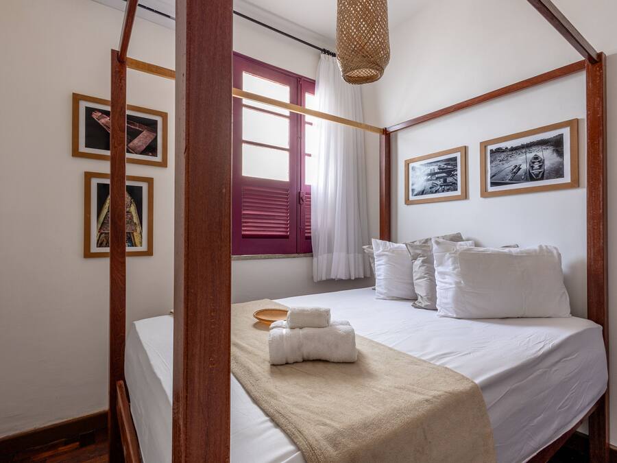 Quarto com cama de dossel, roupa de cama branca e manta bege. Há quadros nas paredes e uma janela com cortinas brancas.