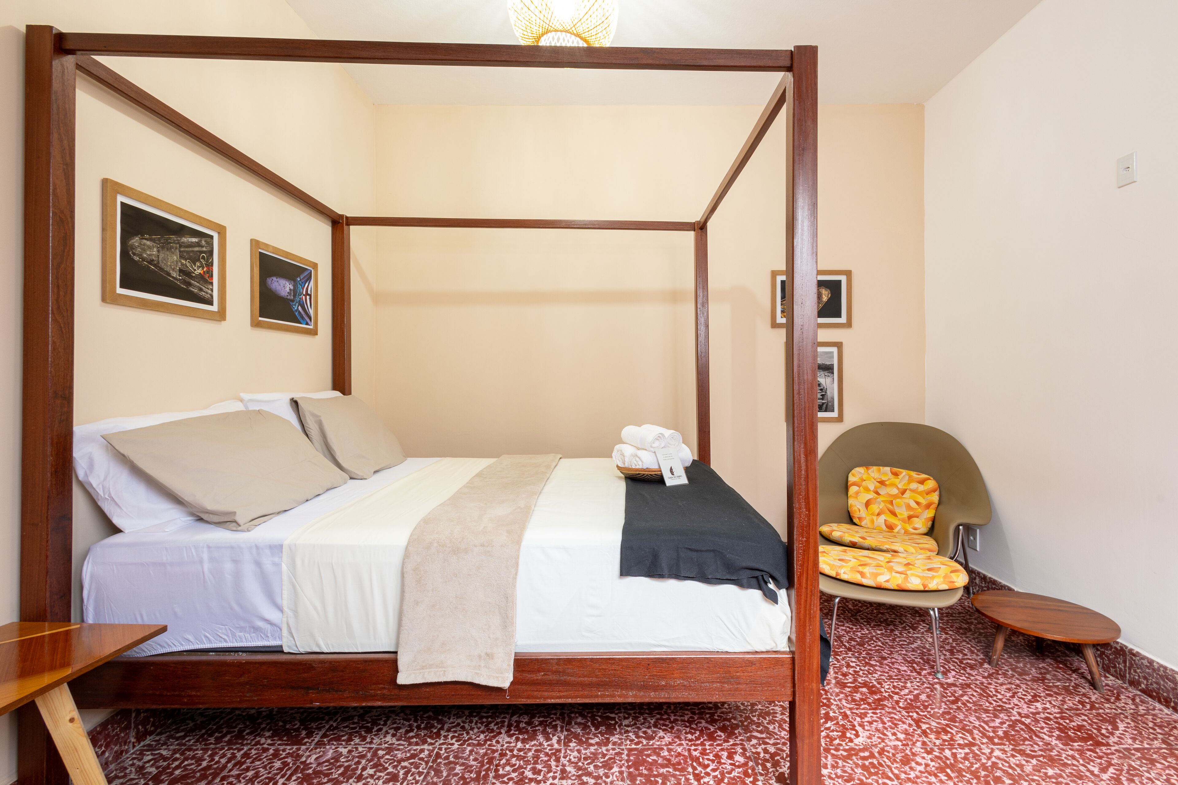 Uma cama de dossel com estrutura de madeira, lençóis brancos e um cobertor bege. Há duas quadros pendurados na parede acima da cama.