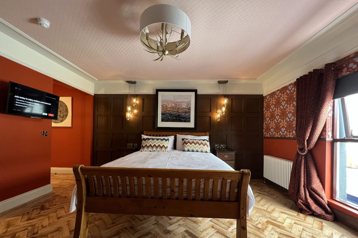 Quarto com cama de madeira, televisão montada na parede, luminária no teto e janela com cortinas.
