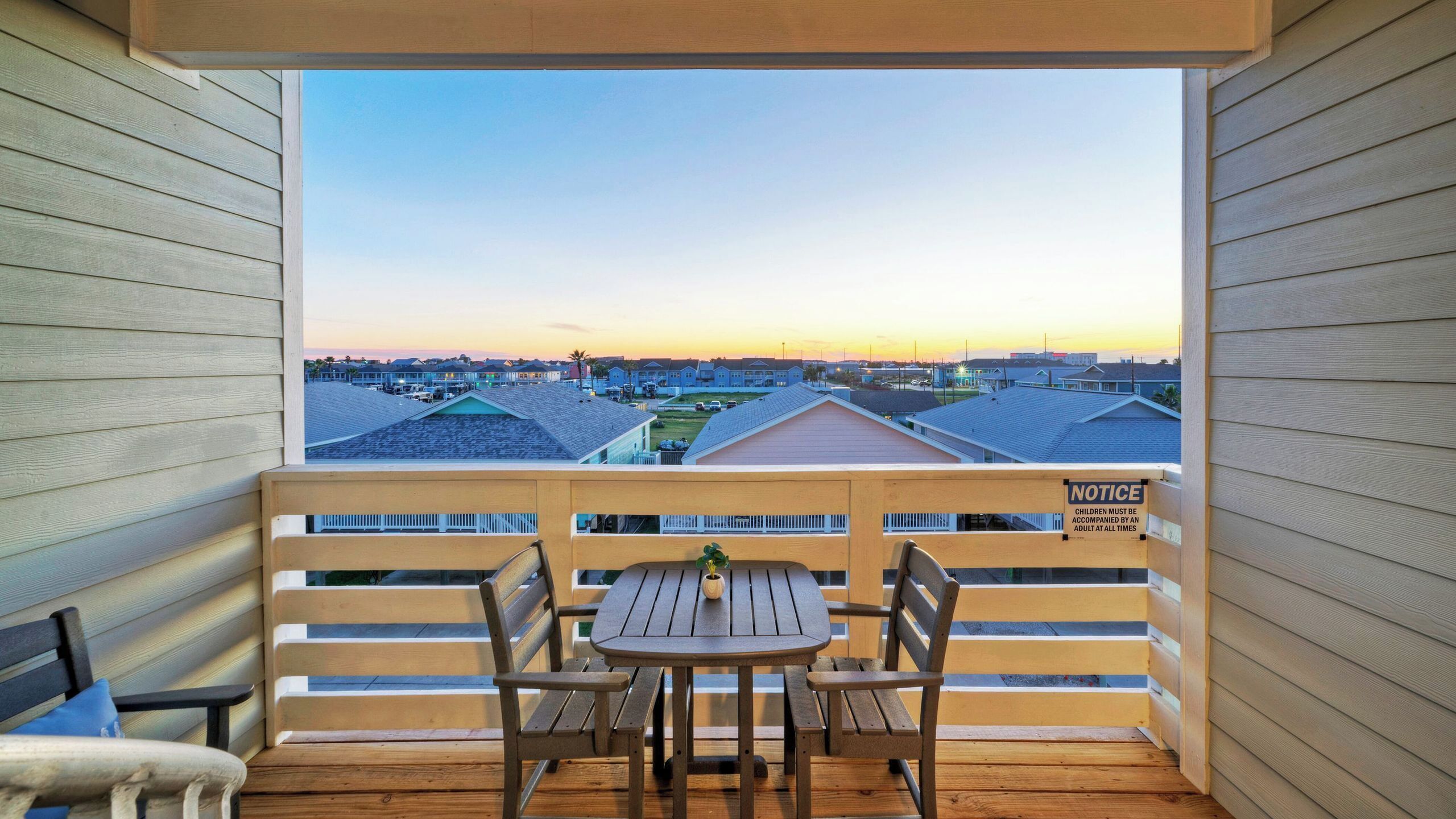 Um deck de madeira com mesa e cadeiras, com vista para uma área residencial com casas e um céu claro.