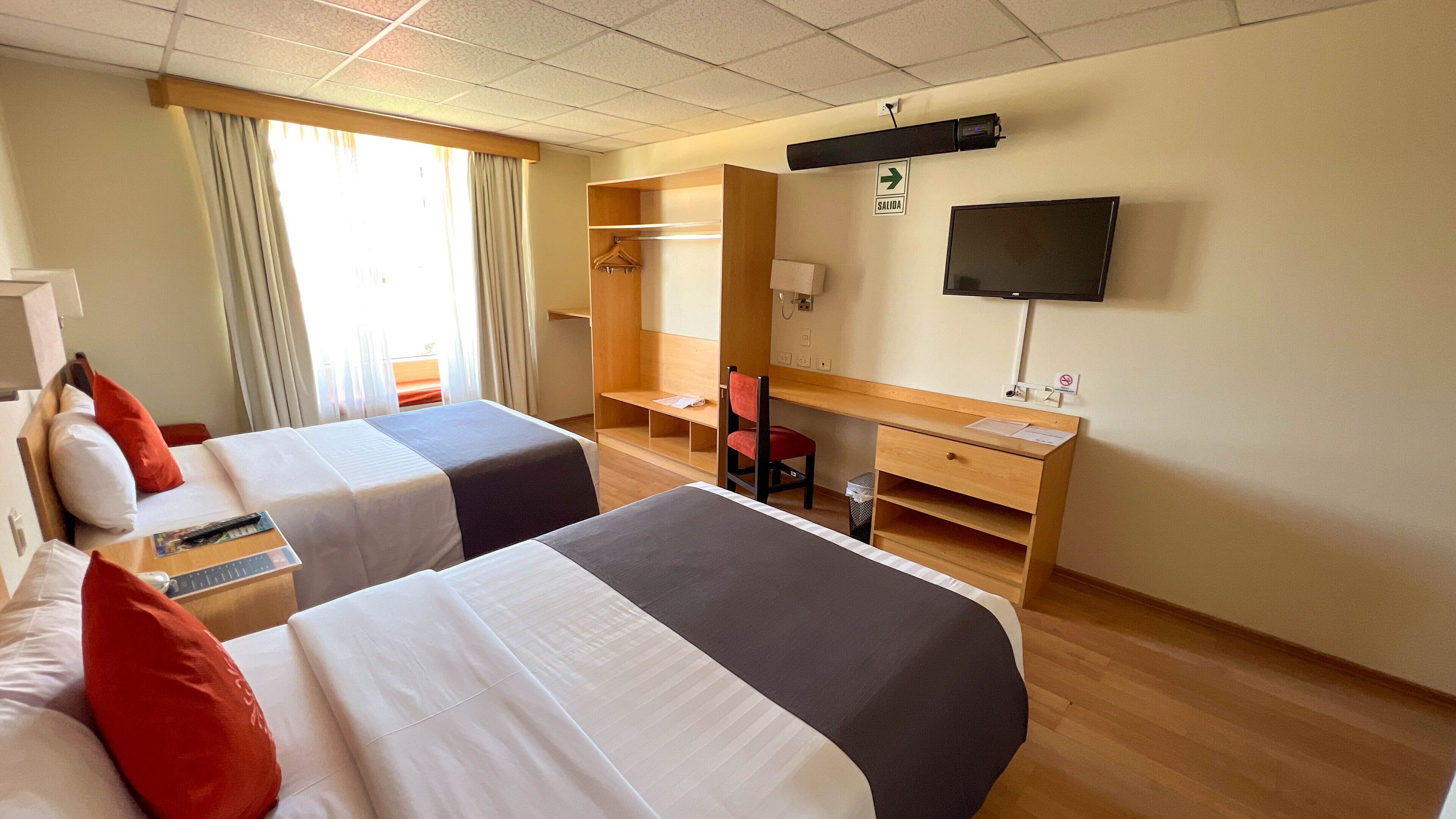 Quarto de hotel com duas camas, uma escrivaninha, televisão e janela com cortinas.