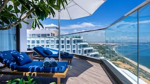 Um terraço com espreguiçadeiras azuis e um grande guarda-sol, com vista para uma paisagem costeira.