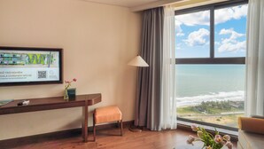 Quarto de hotel com uma grande janela com vista para o oceano, uma escrivaninha com abajur e um mapa emoldurado na parede.