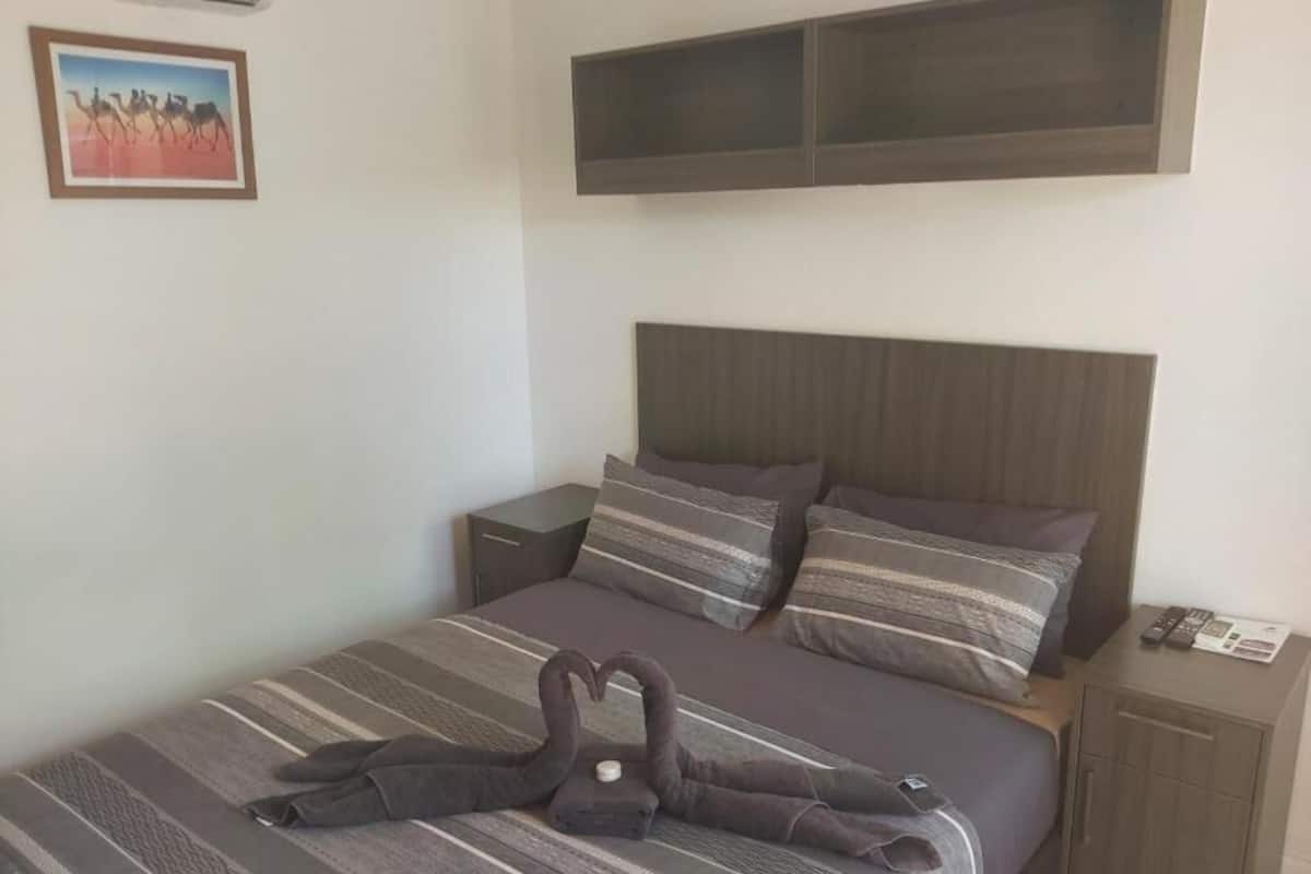 Uma cama bem arrumada com roupa de cama listrada, arranjo de travesseiros em formato de coração, mesa de cabeceira com controle remoto e um quadro na parede.