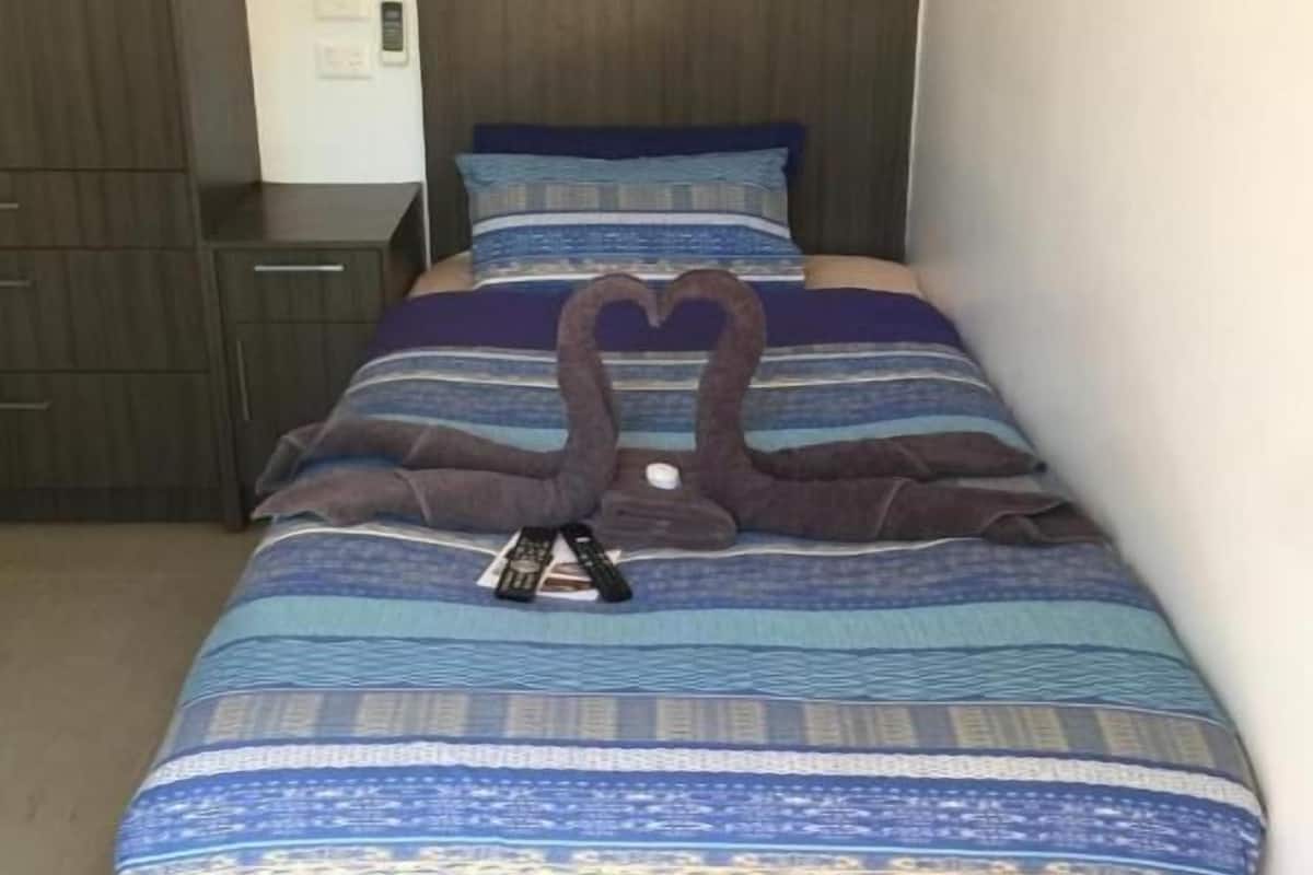 Uma cama de solteiro com colcha listrada, arranjo de almofadas em formato de coração e um controle remoto sobre a cama.