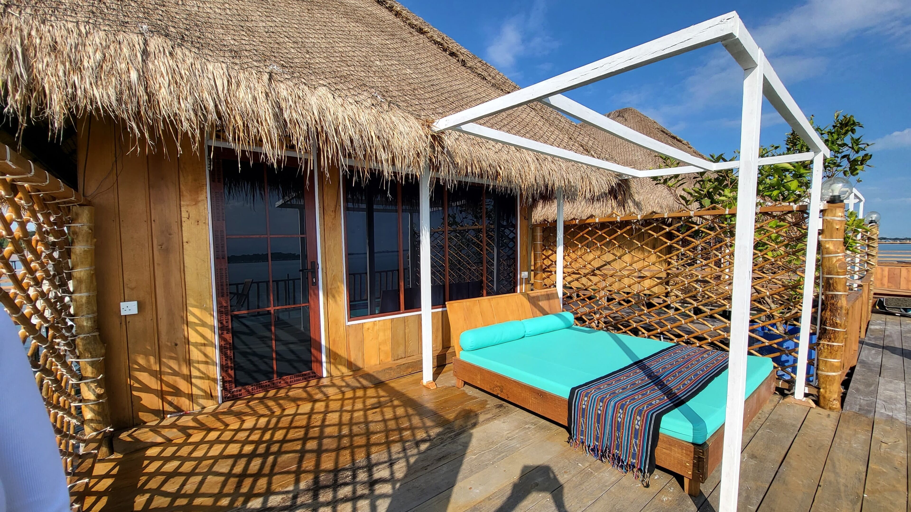 Uma cabana de madeira com telhado de palha, um deck de madeira e uma área de estar coberta com um almofada azul.