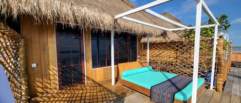 Uma cabana de madeira com telhado de palha, um deck de madeira e uma área de estar coberta com um almofada azul.
