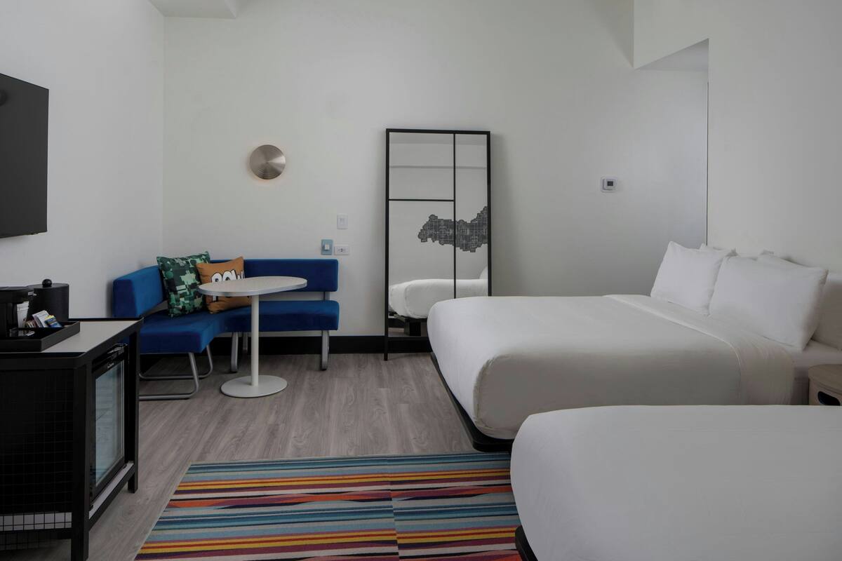Quarto de hotel com cama, escrivaninha com computador, mesinha pequena, sofá e tapete listrado e colorido.