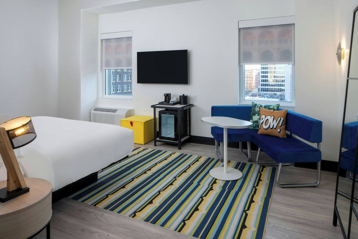 Quarto de hotel com cama, escrivaninha, cadeira, televisão e janela com vista para a cidade.