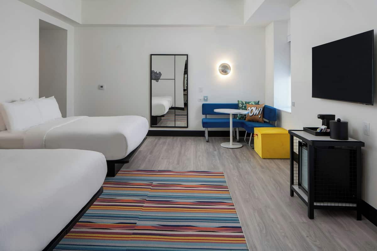 Quarto de hotel com duas camas, um espelho, uma mesa pequena com abajur, uma televisão e um tapete listrado e colorido.