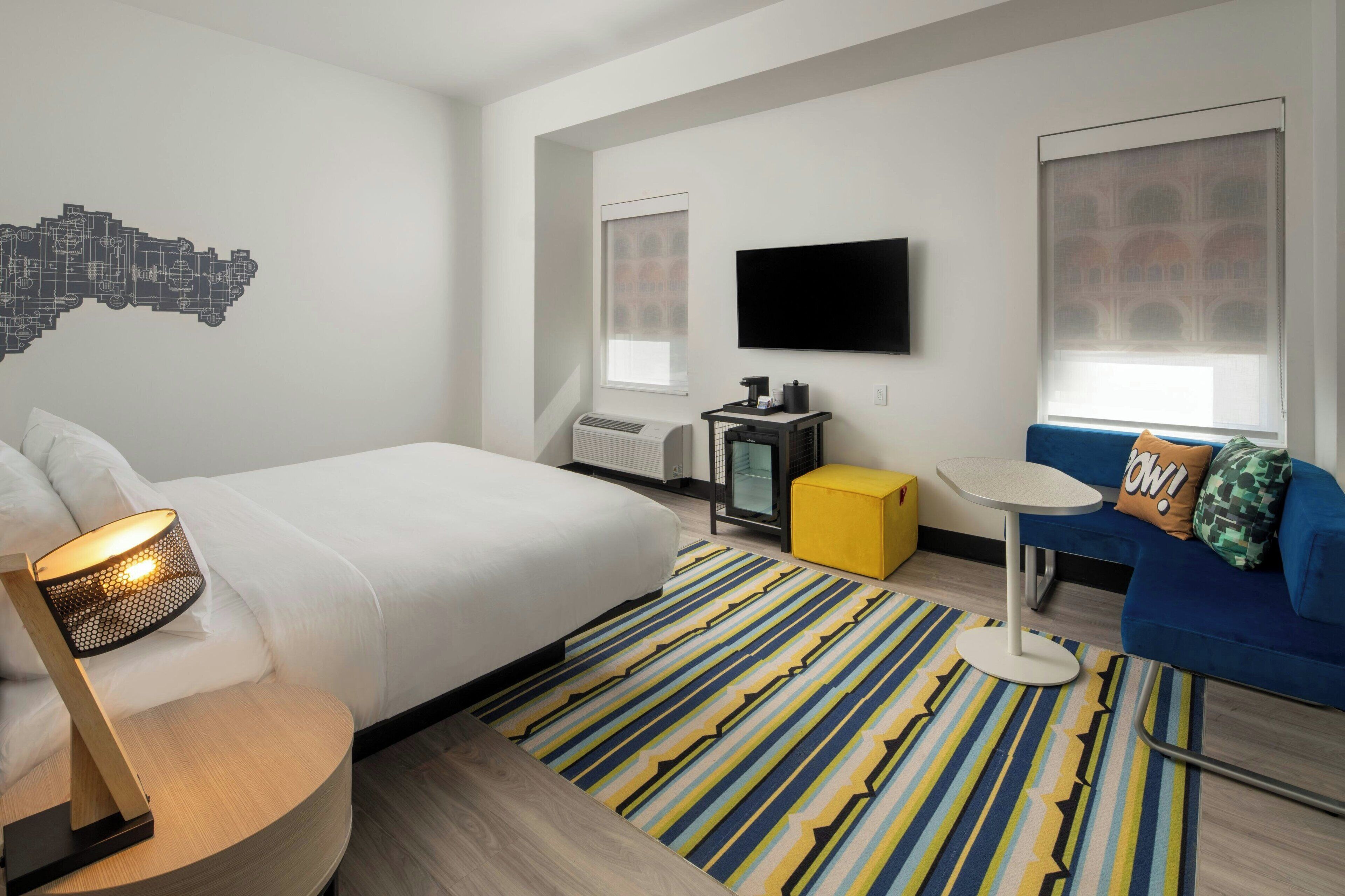 Quarto de hotel moderno com uma cama grande, um sofá pequeno, uma televisão e uma mesa de centro.