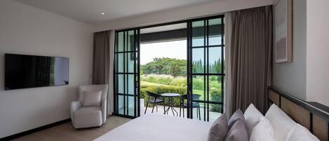 Quarto de hotel moderno com uma cama grande, televisão de tela plana, área de estar com mesa e cadeiras, e varanda com vista para área verde.