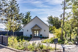 Uma casa pequena e branca, térrea, com telhado em duas águas, cercada por um jardim com diversas plantas e árvores.