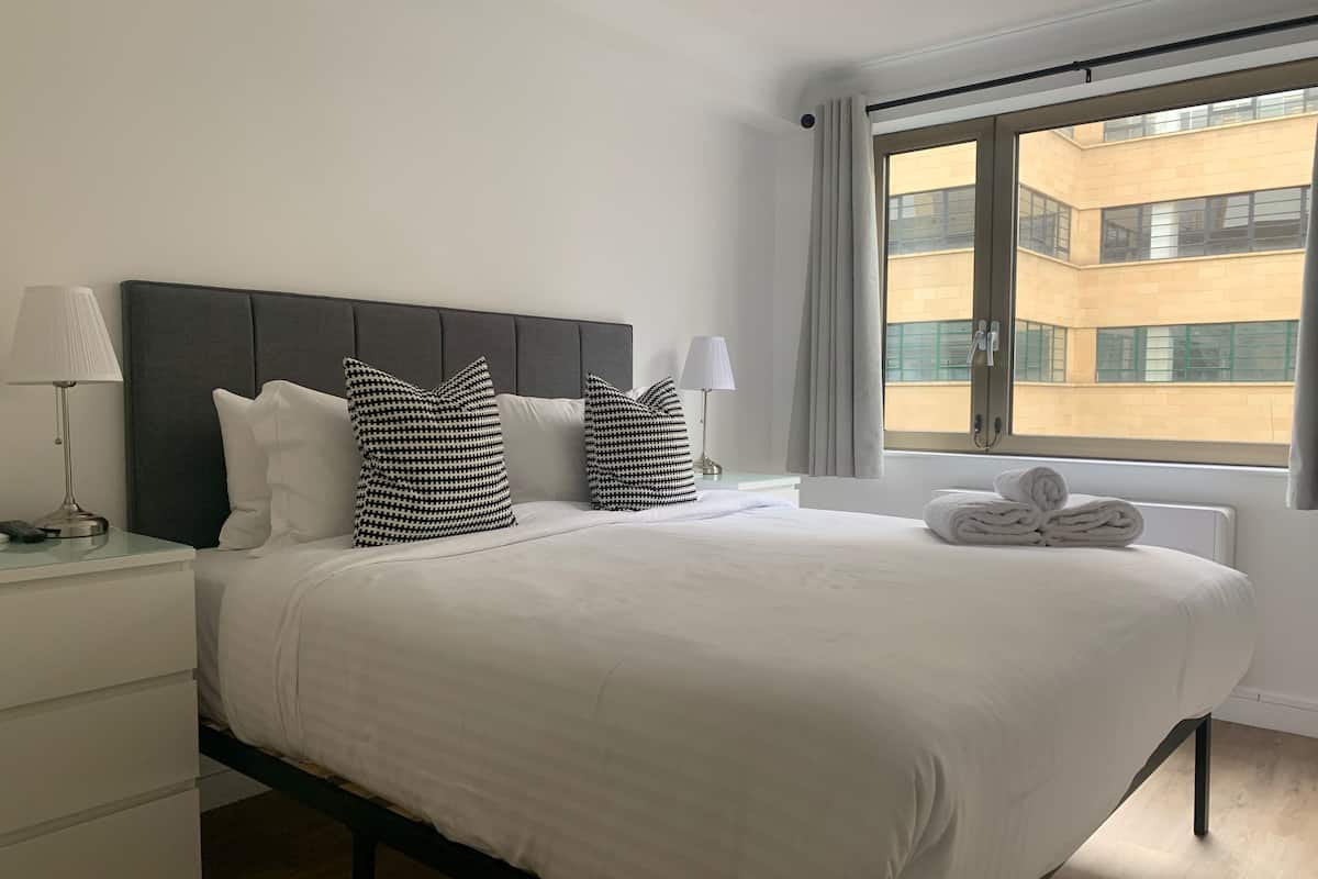 Quarto de hotel com cama, duas luminárias de cabeceira, janela com cortinas e vista para um edifício.