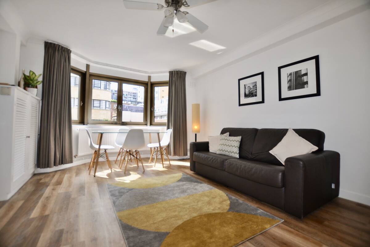 Uma sala de estar moderna com um sofá marrom escuro, uma mesa de jantar com cadeiras e uma janela grande com cortinas.