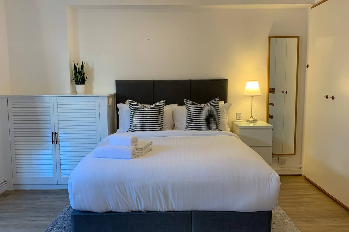 Cama bem arrumada com cabeceira escura, roupa de cama branca e duas almofadas estampadas. Tem uma pequena mesa de cabeceira com luminária, um vaso de planta e um espelho na parede.