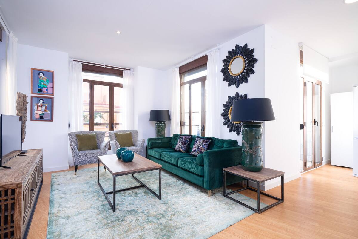 Uma sala de estar com um sofá verde, uma mesa de centro de madeira, uma poltrona estampada, um vaso azul e um espelho grande na parede.