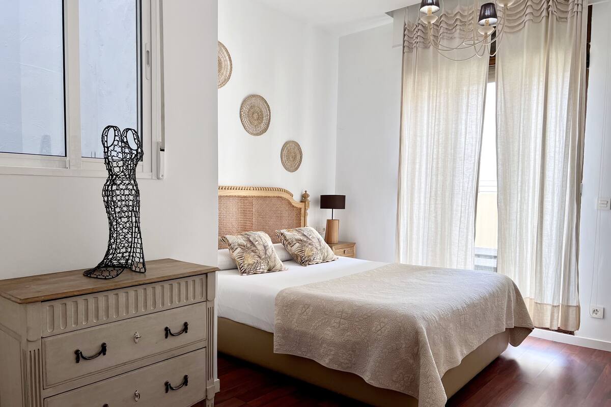 Quarto com cômoda de madeira, cama com colcha bege, mesa de cabeceira com abajur e arte decorativa na parede.