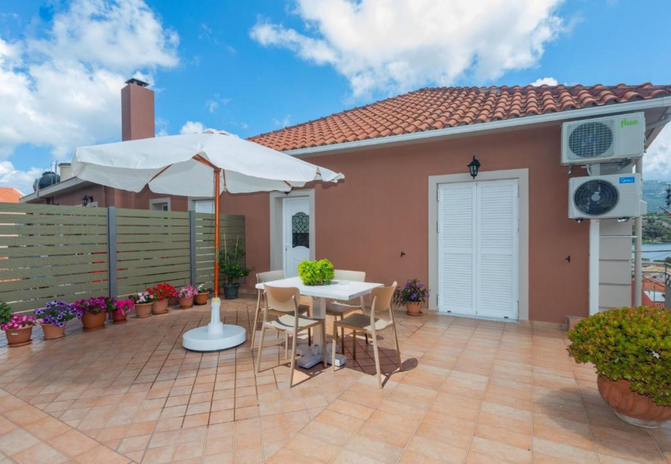 Pátio externo com mesa e cadeiras, cercado por plantas em vasos e um guarda-sol branco.