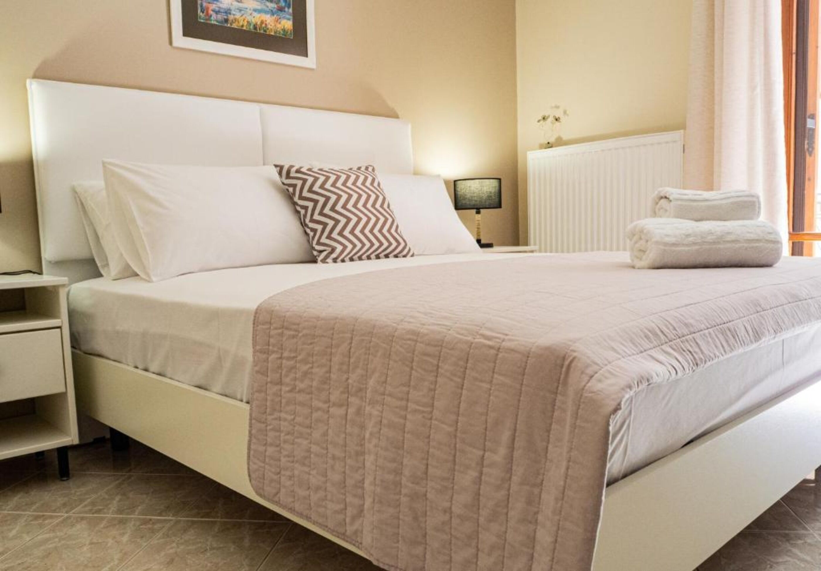 Uma cama bem arrumada com cobertura acolchoada, uma mesinha de cabeceira com abajur e um radiador ao fundo.