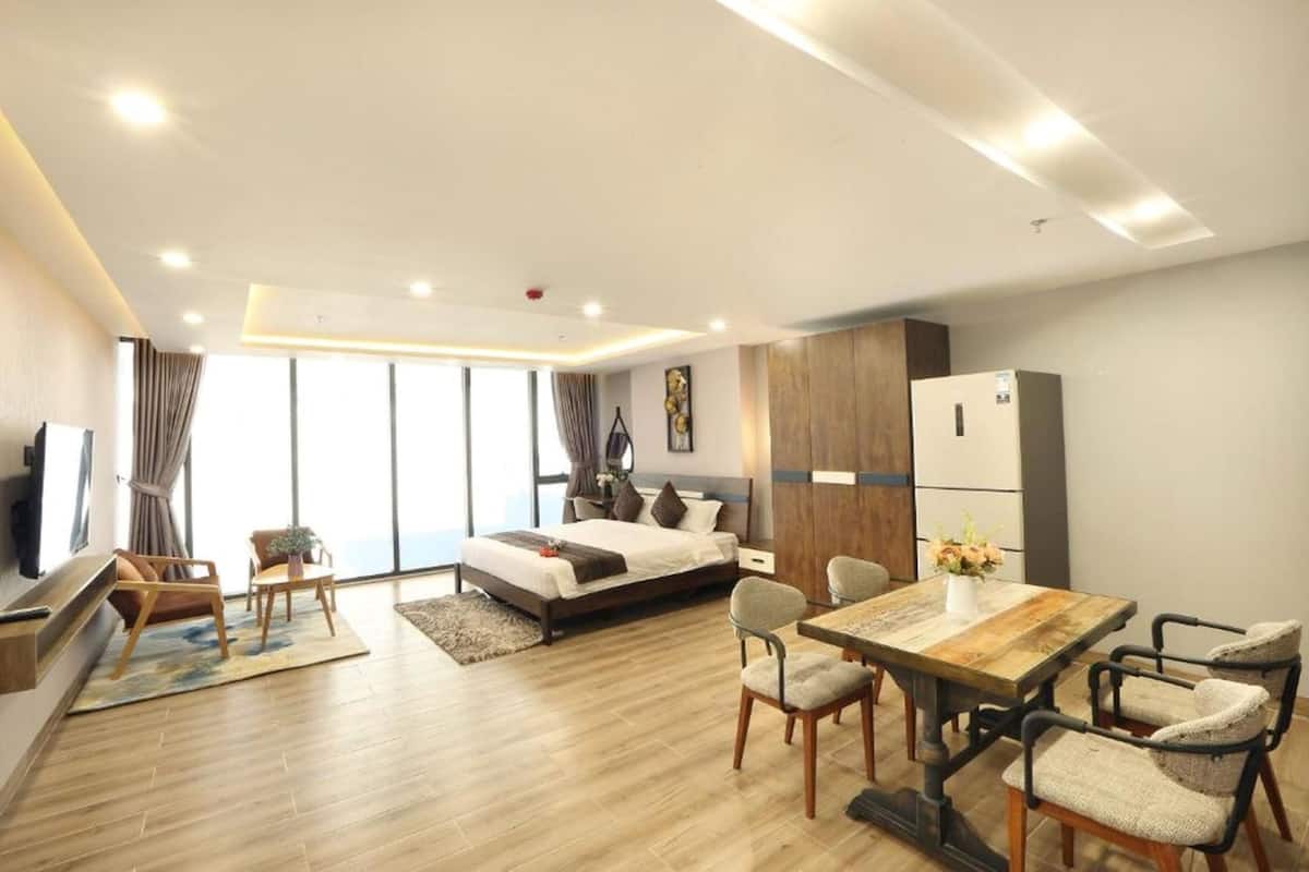 Quarto de hotel moderno com uma cama grande, área de jantar com mesa e cadeiras de madeira, TV e uma janela ampla com vista.