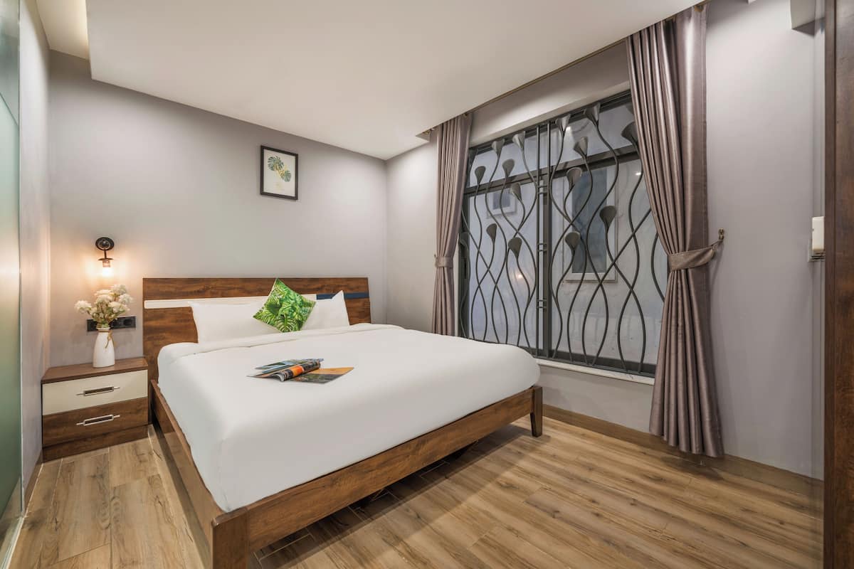 Um quarto bem arrumado com uma cama de madeira, uma mesa de cabeceira com um vaso de flores, uma janela com cortinas decorativas e um aparelho de luz fixado na parede.