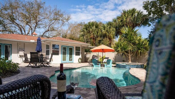 Área da piscina com espreguiçadeiras, uma mesa com uma garrafa de vinho e uma casa ao fundo.
