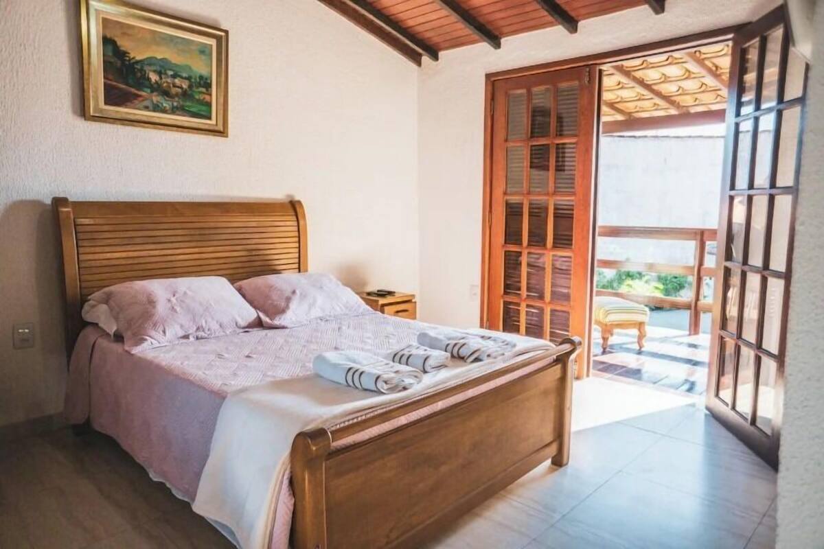 Quarto com cama de madeira, mesa de cabeceira, quadro na parede e porta deslizante para área externa.