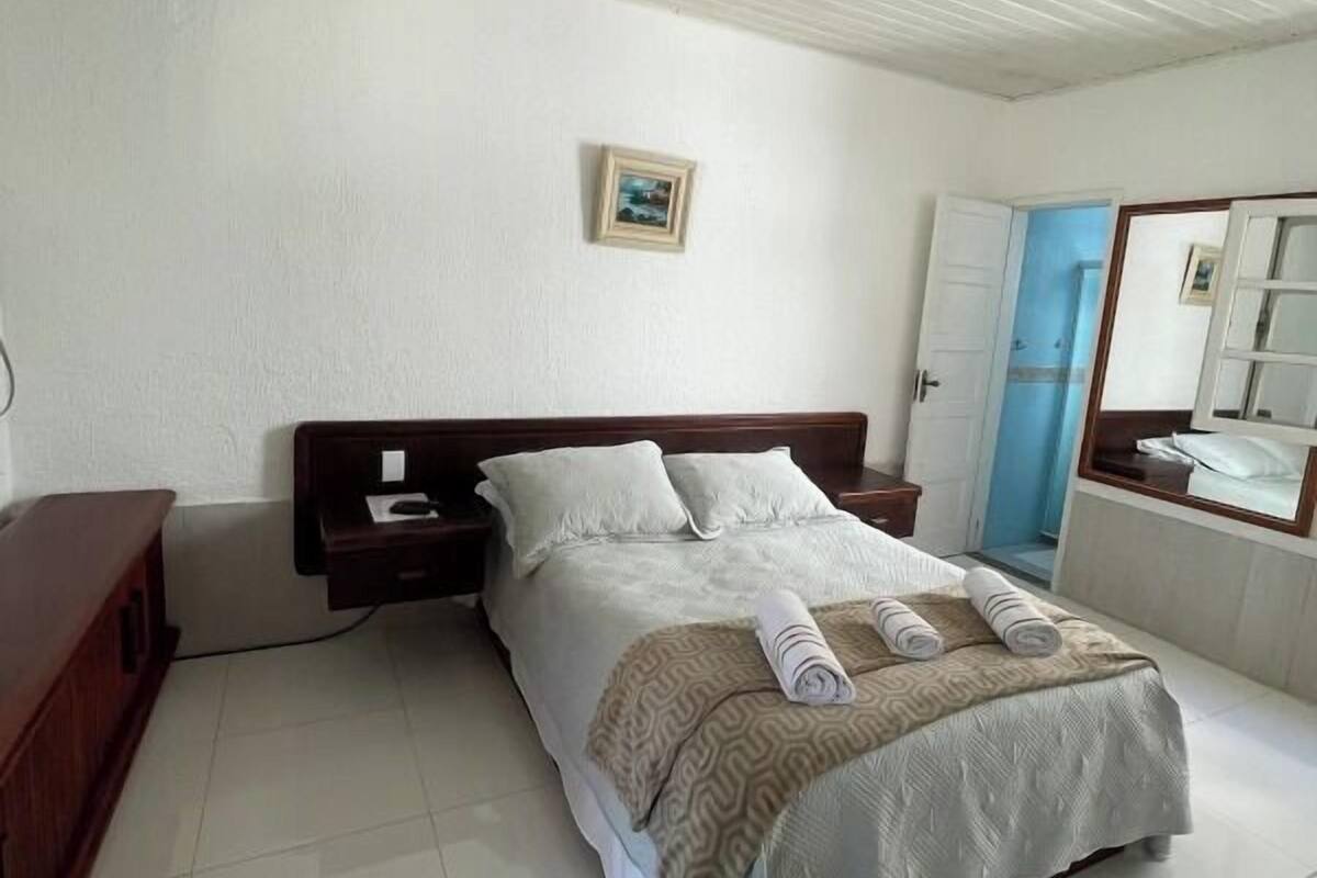 Quarto de hotel limpo e bem iluminado com uma cama grande, mesa de cabeceira, cómoda de madeira e casa de banho com espelho, além de ventoinha no teto.