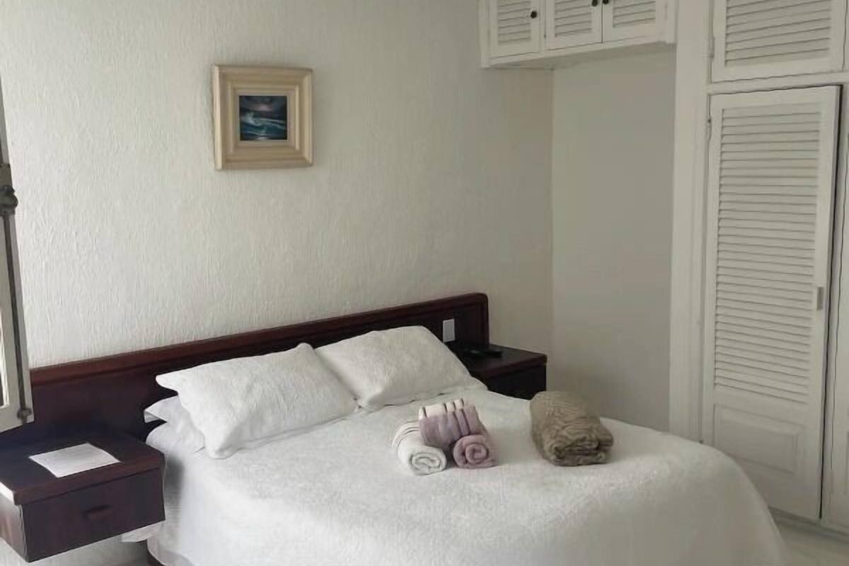 Uma cama bem arrumada com lençóis brancos, cabeceira de madeira e mesa de cabeceira com abajur.