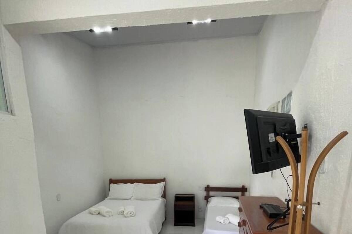 Quarto pequeno e limpo com duas camas, cômoda de madeira, televisão e aquecedor.