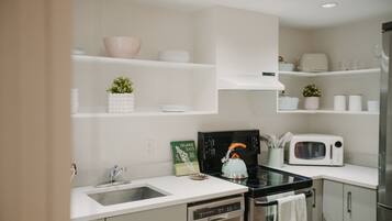 Uma cozinha moderna com bancadas brancas, lava-louças, fogão, micro-ondas e prateleiras com louças e plantas em vasos.