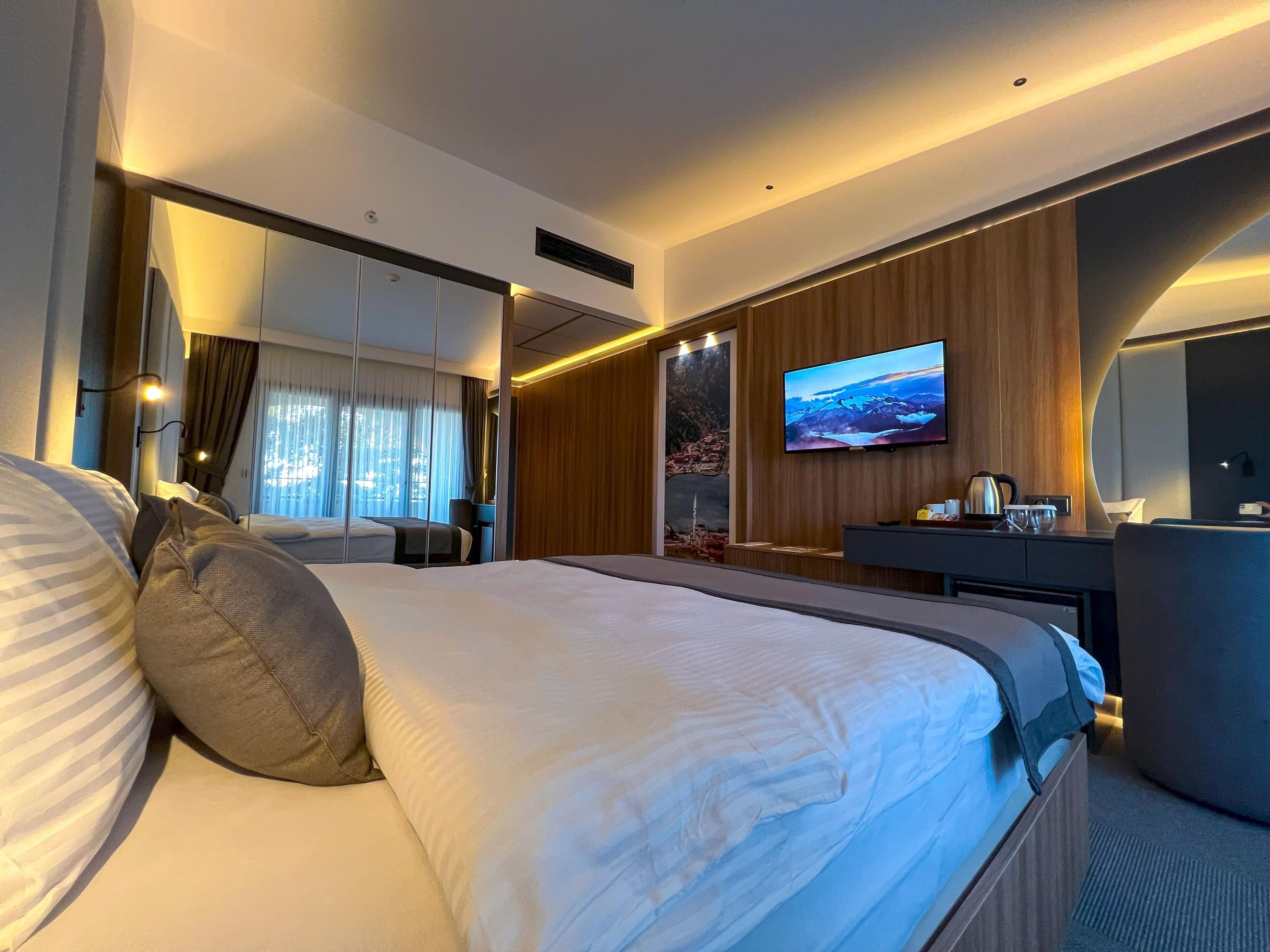 Quarto de hotel moderno com uma cama grande, uma TV de tela plana, uma escrivaninha com cadeira e uma janela ampla com cortinas.