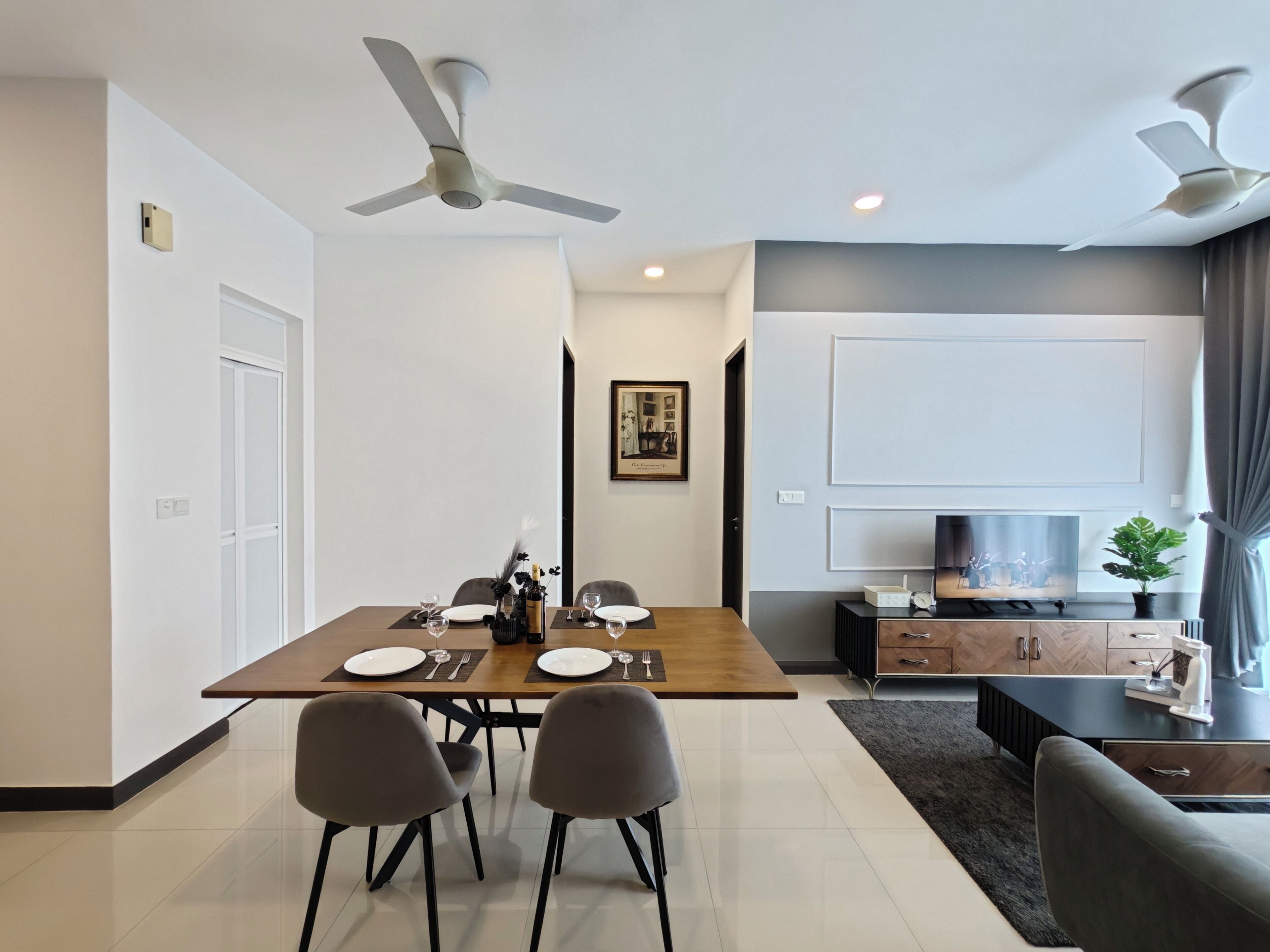 Uma sala de estar moderna com área de jantar, apresentando uma mesa de jantar de madeira para duas pessoas, duas cadeiras cinzas, um sofá preto, uma televisão e ventiladores de teto.